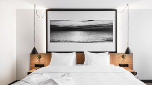 Quarto de hotel moderno com uma cama grande, roupa de cama branca, duas mesas de cabeceira com luminárias e uma fotografia emoldurada de uma praia na parede.