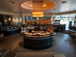 Uma área de buffet com ilha central, disposição de mesas e um mural na parede.