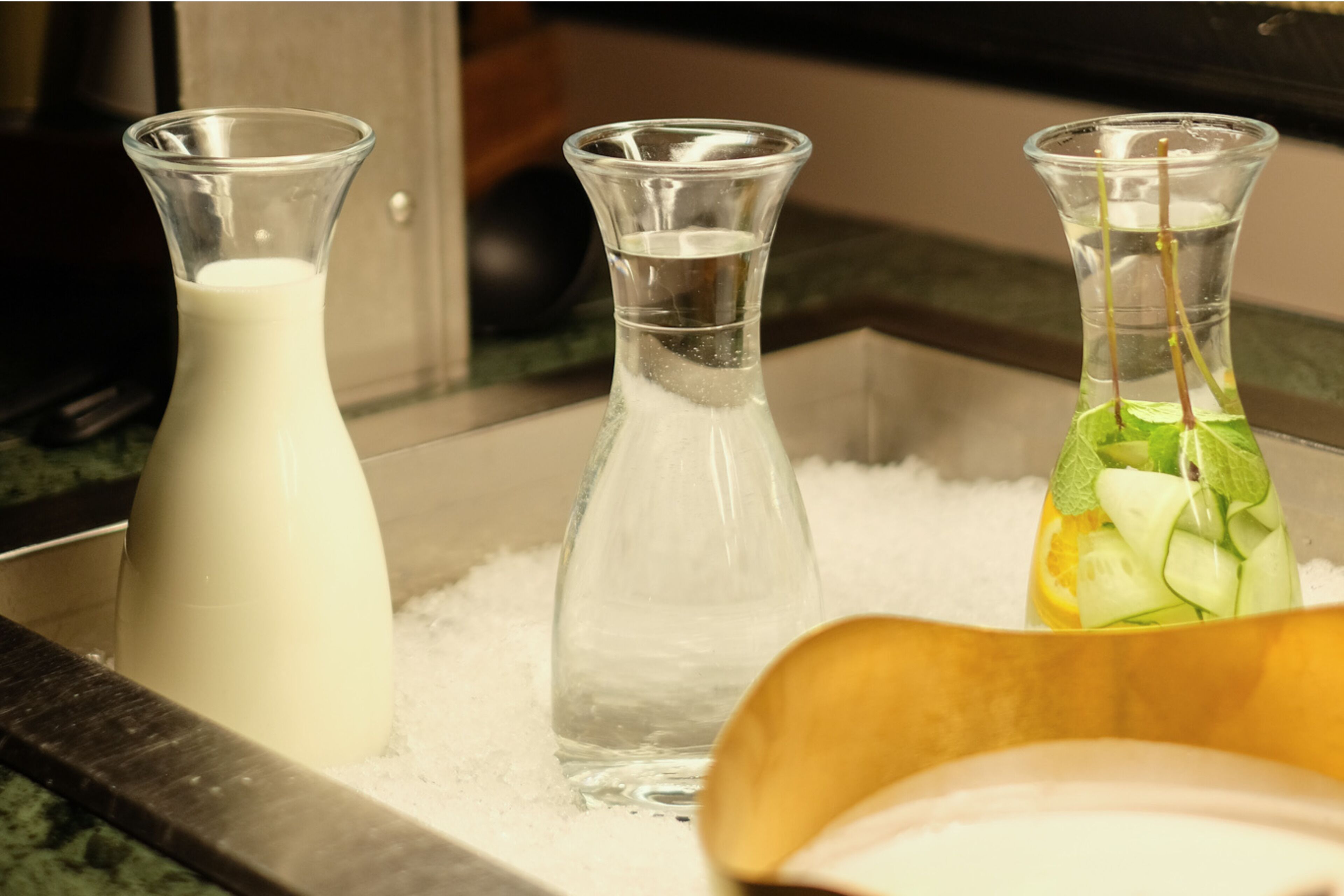 Três vasos de vidro sobre uma bandeja com gelo, um contendo um líquido branco e outro com uma bebida verde e folhas de hortelã.
