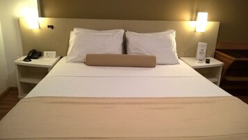 Quarto de hotel com uma cama bem arrumada, duas mesas de cabeceira, uma luminária, um telefone e uma placa com o nome do quarto.