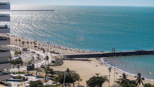 Uma praia costeira com calçadão, palmeiras e um cais.