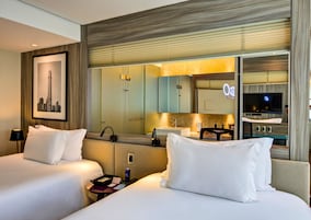 Quarto de hotel com duas camas, um espelho grande, uma televisão e uma fotografia da paisagem urbana na parede.