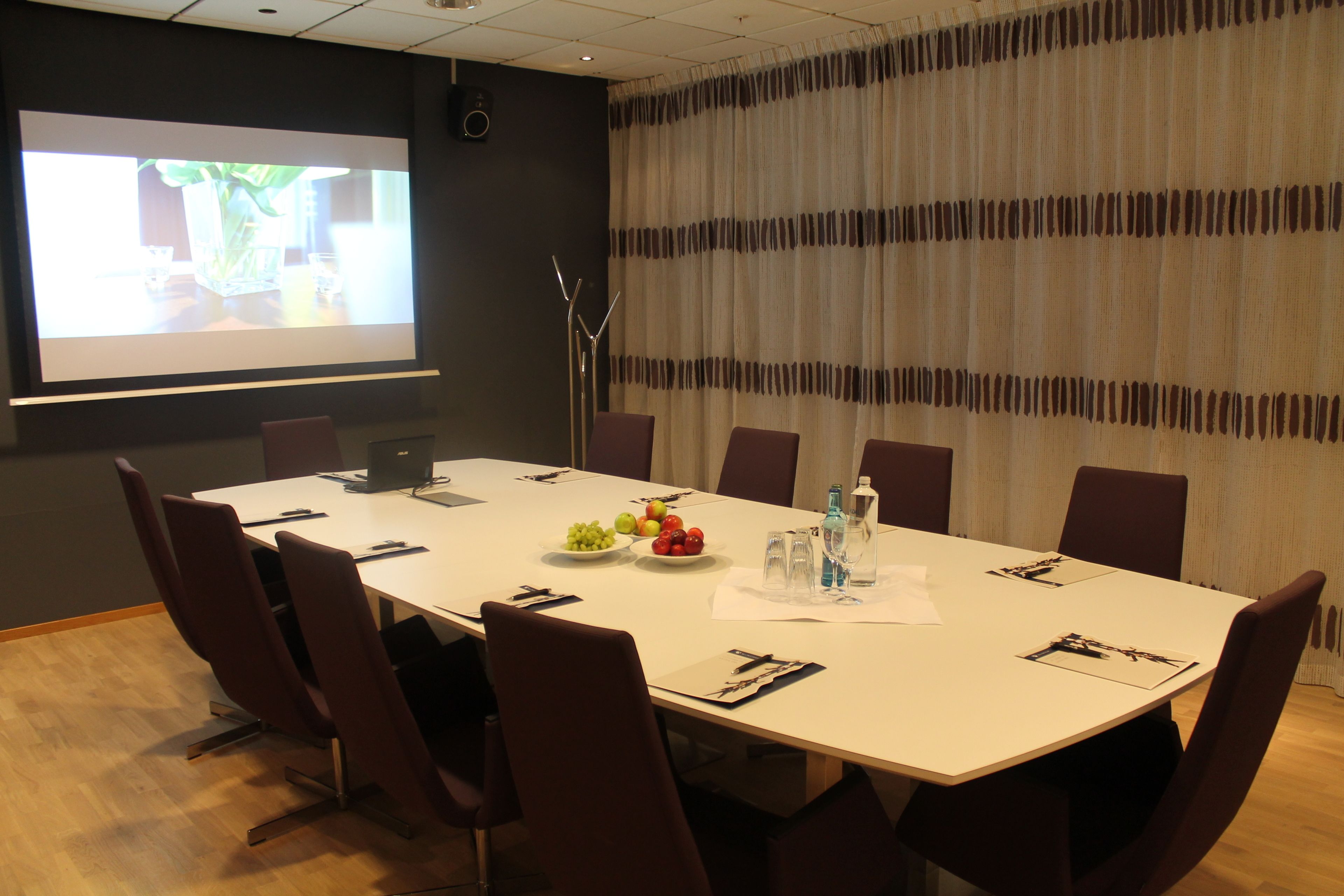 Uma sala de conferências com uma mesa comprida, cadeiras, tela de projetor e uma planta.