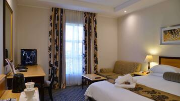 Quarto de hotel com cama, escrivaninha com cafeteira, cadeira, televisão e janela com cortinas.