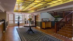 Um hall espaçoso com piso de azulejos, uma mesa redonda central com um vaso de flores, uma escada com corrimãos de madeira e uma grande janela com vista para o exterior.