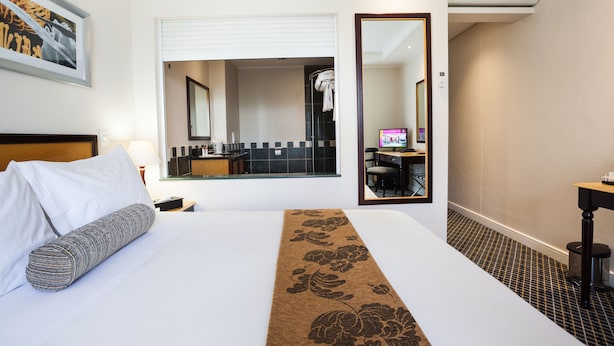 Quarto de hotel com cama, mesa de cabeceira, luminária, espelho e vista para o banheiro e cozinha através de uma janela.