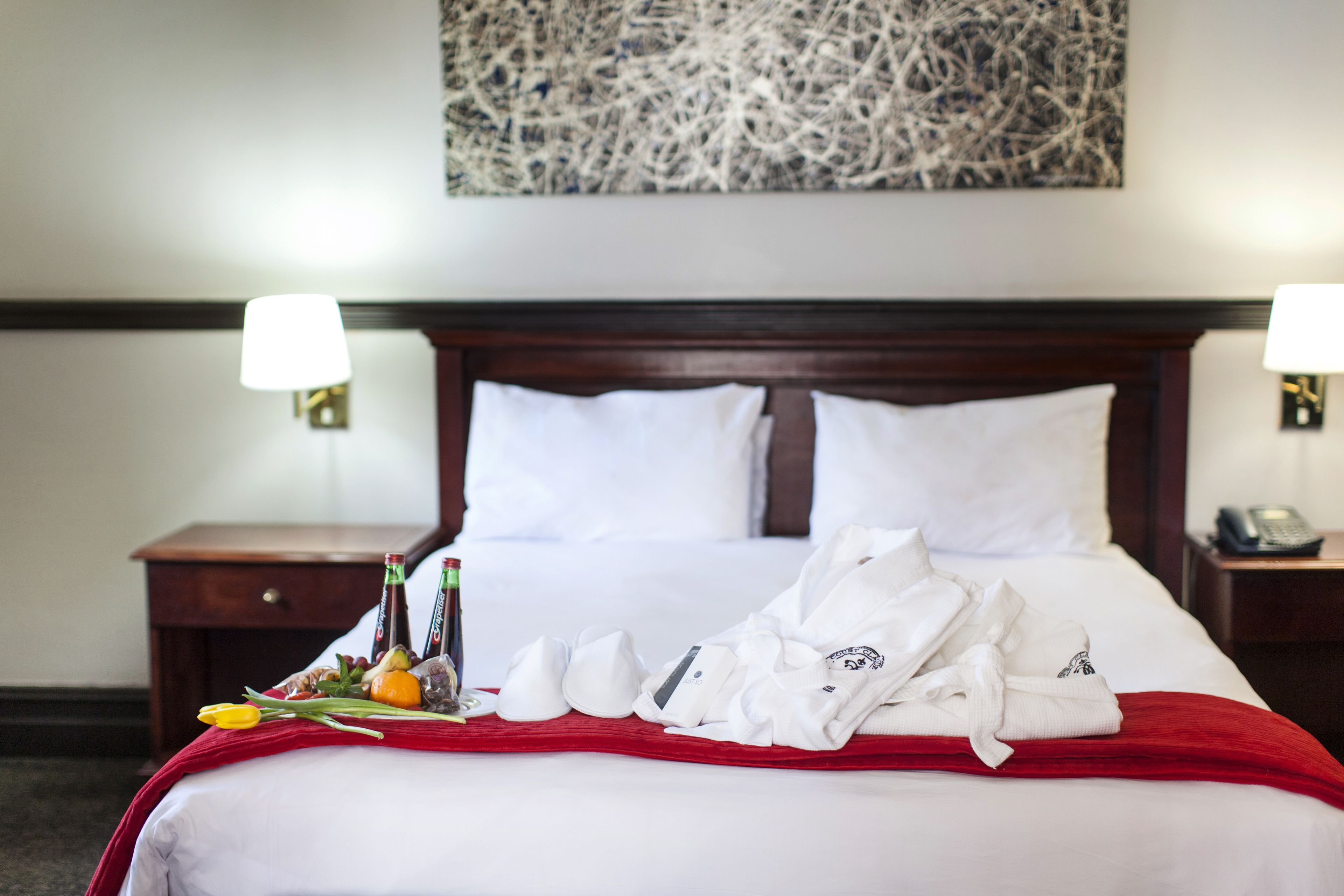 Quarto de hotel com cama bem arrumada, manta vermelha e roupão sobre a cama. Há uma mesa de cabeceira com uma luminária e uma garrafa de água.