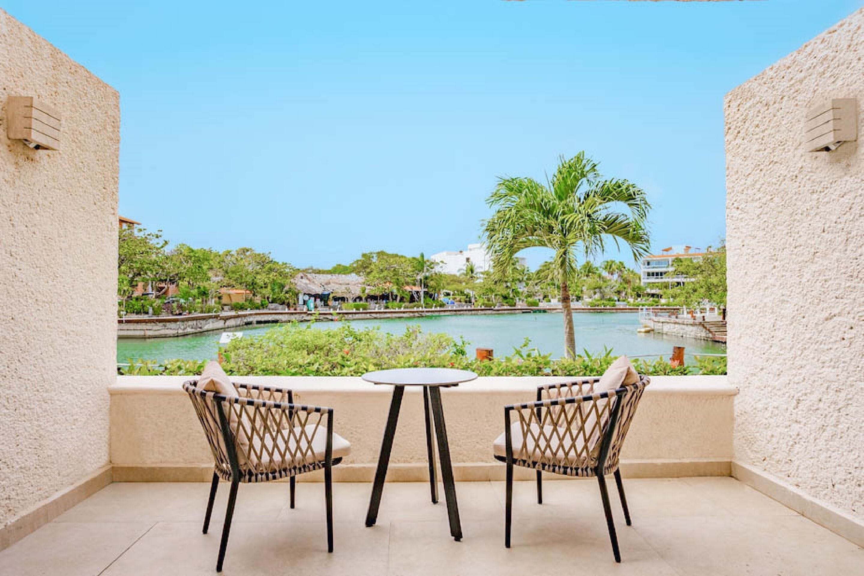 Varanda com duas cadeiras de vime e uma mesa pequena, com vista para uma piscina e vegetação.