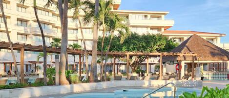 Um hotel com piscina, palmeiras e uma construção com telhado de palha.