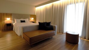 Quarto de hotel moderno com uma cama grande, um sofá, um banquinho redondo e uma mesa de cabeceira com abajur.