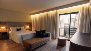 Quarto de hotel moderno com uma cama grande, um sofá, um banquinho redondo pequeno e uma varanda com cortinas.