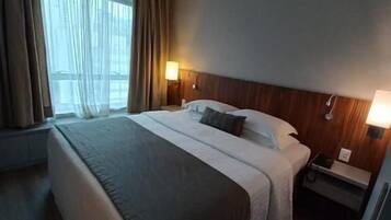Quarto de hotel com uma cama grande, uma mesinha de cabeceira com abajur e uma janela com cortinas.