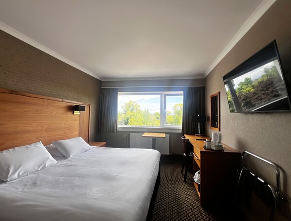 Quarto de hotel com uma cama grande, uma escrivaninha, uma cadeira, uma televisão e uma janela com vista para árvores.