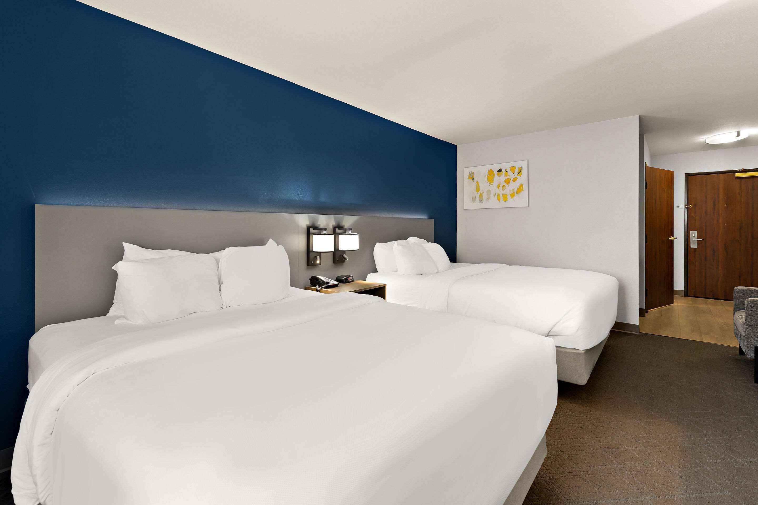 Quarto de hotel com duas camas, uma mesinha de cabeceira com abajur, uma porta de madeira e uma pequena área de estar.