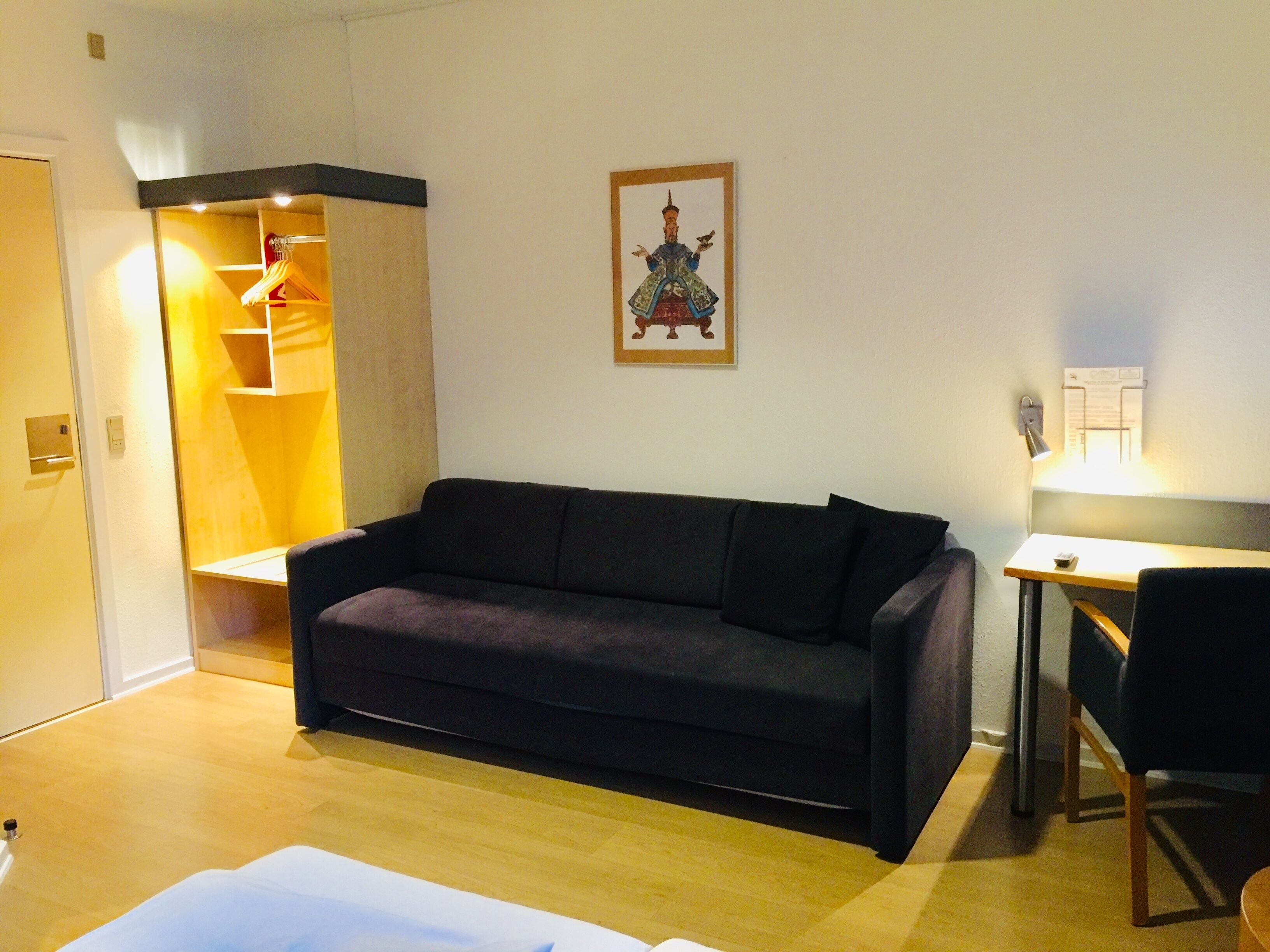 Quarto com um sofá de cor escura, uma secretária pequena com uma cadeira, um armário de madeira e um quadro na parede.