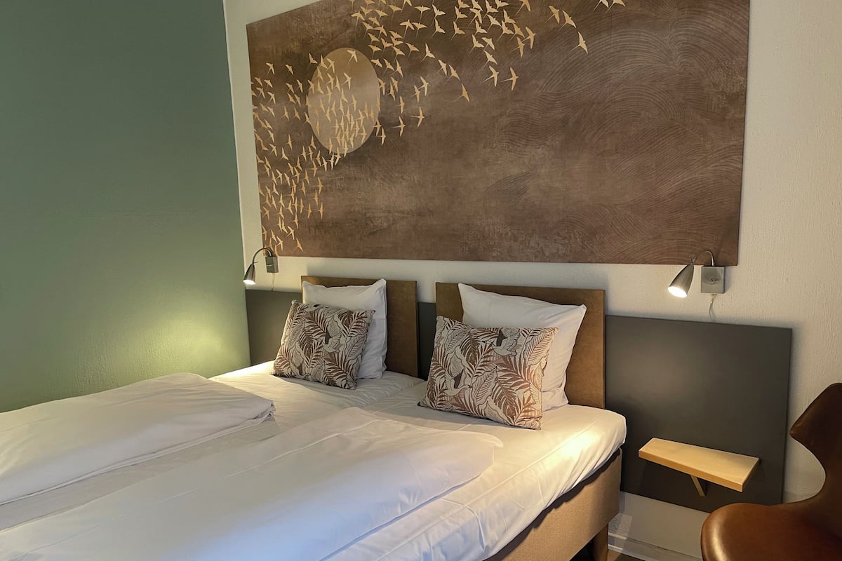 Cama bem arrumada com lençóis brancos e duas almofadas estampadas, cabeceira de madeira, mesa de cabeceira com abajur e uma obra de arte na parede acima da cama.