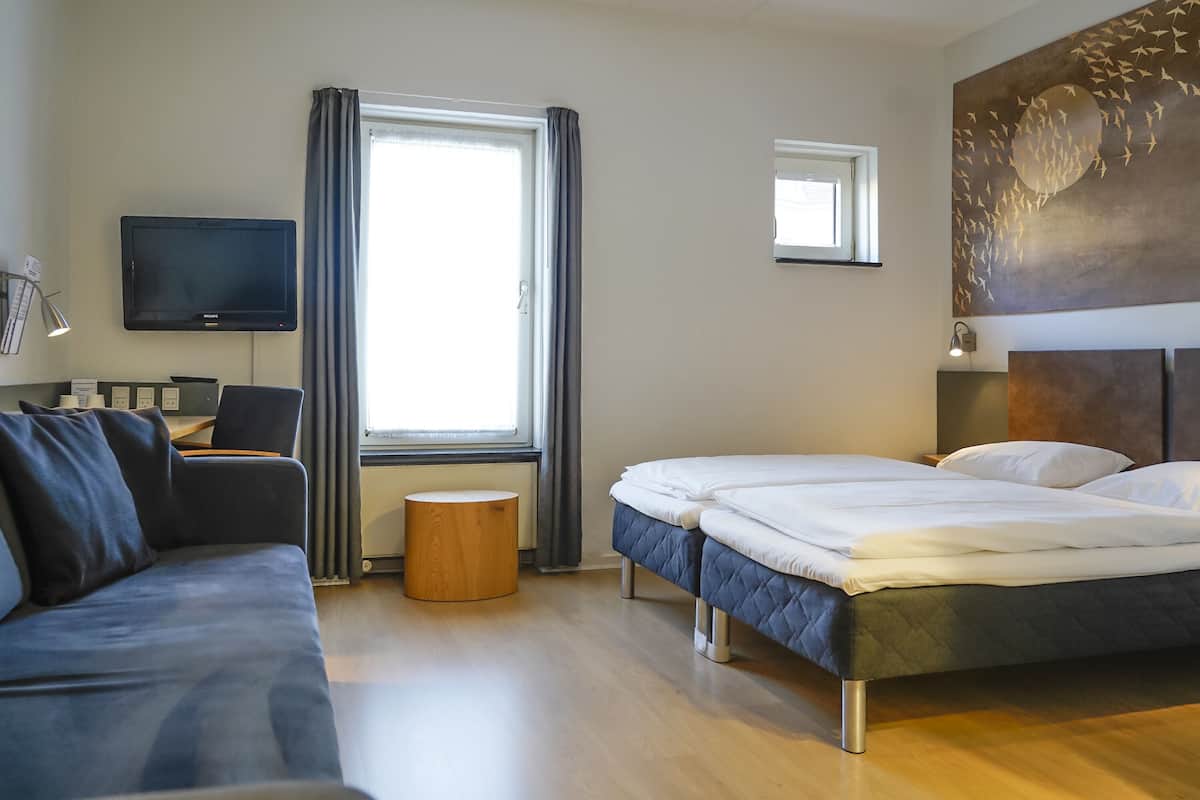 Quarto de hotel com uma cama, sofá, televisão e janela com cortinas.