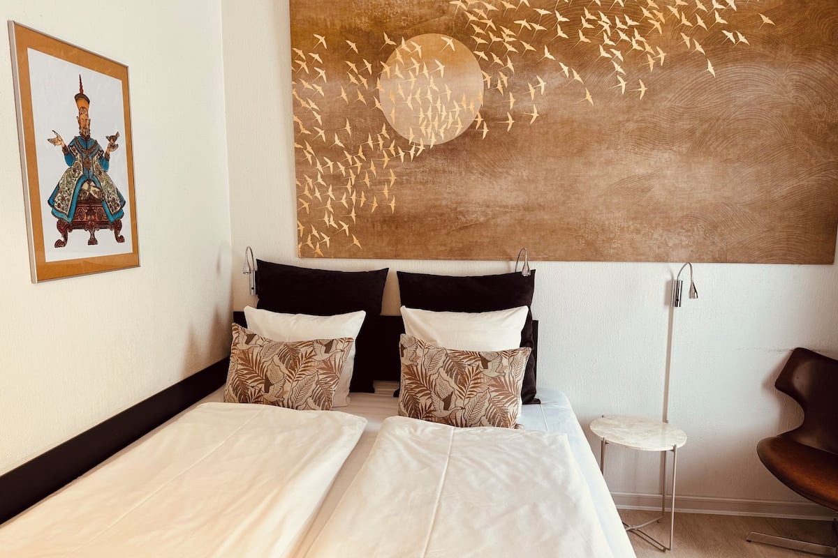 Quarto de hotel com duas camas, uma obra de arte de aves fixada na parede, uma mesinha e uma cadeira.