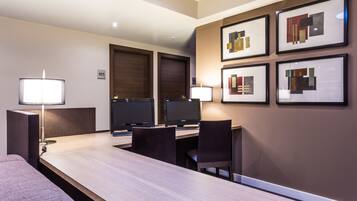 Quarto de hotel com uma longa escrivaninha de madeira, dois monitores de computador, duas cadeiras, uma luminária e obras de arte emolduradas na parede.