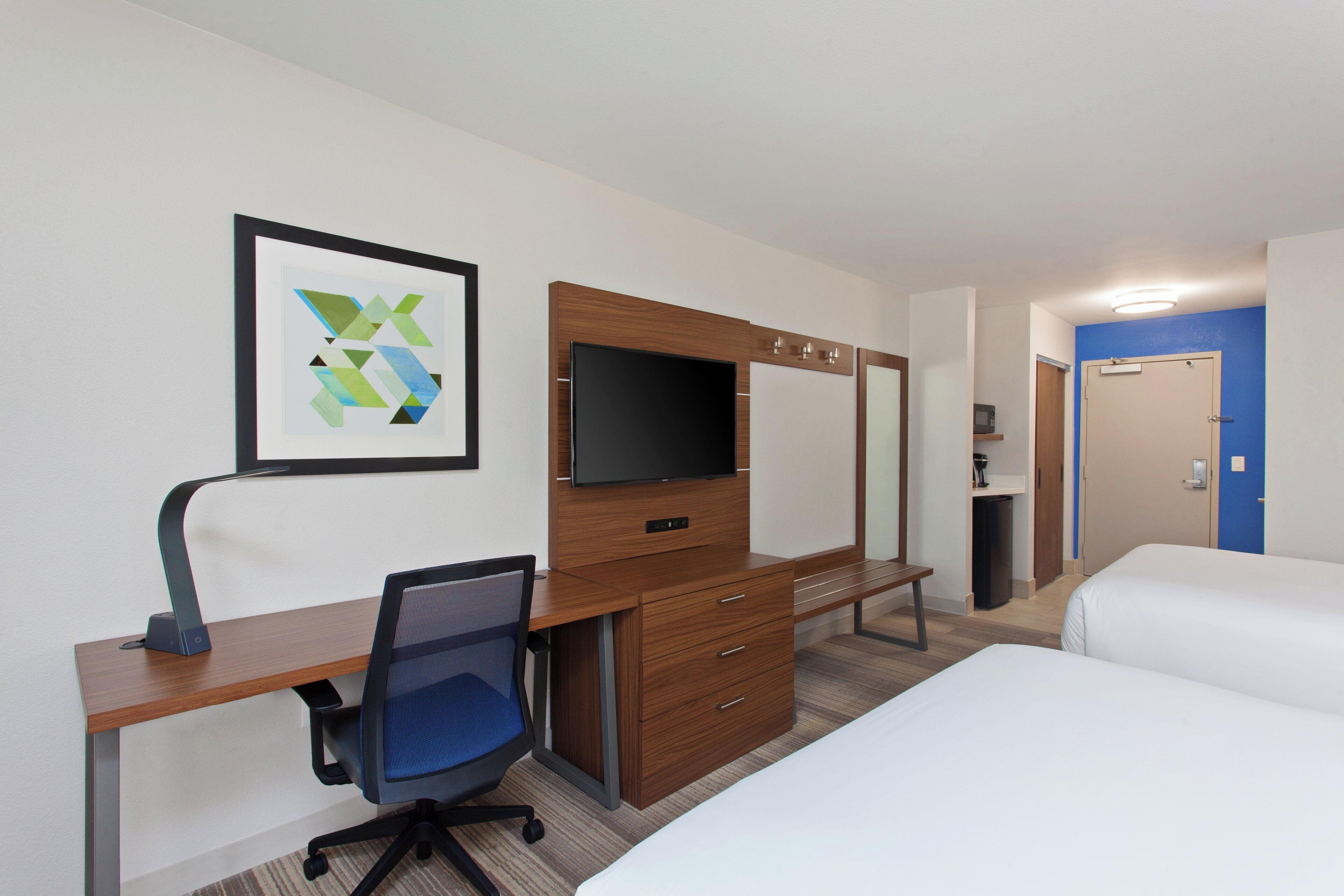 Quarto de hotel com cama, escrivaninha, cadeira, televisão, guarda-roupa e um quadro na parede.