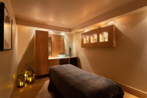 Uma sala de spa moderna com piso de madeira, uma mesa de massagem, um armário de madeira com iluminação e um espelho grande.
