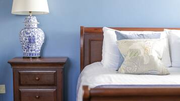 Quarto com mesa de cabeceira de madeira, abajur em vaso azul e branco, cama com roupa de cama branca e almofadas florais, e parede azul.