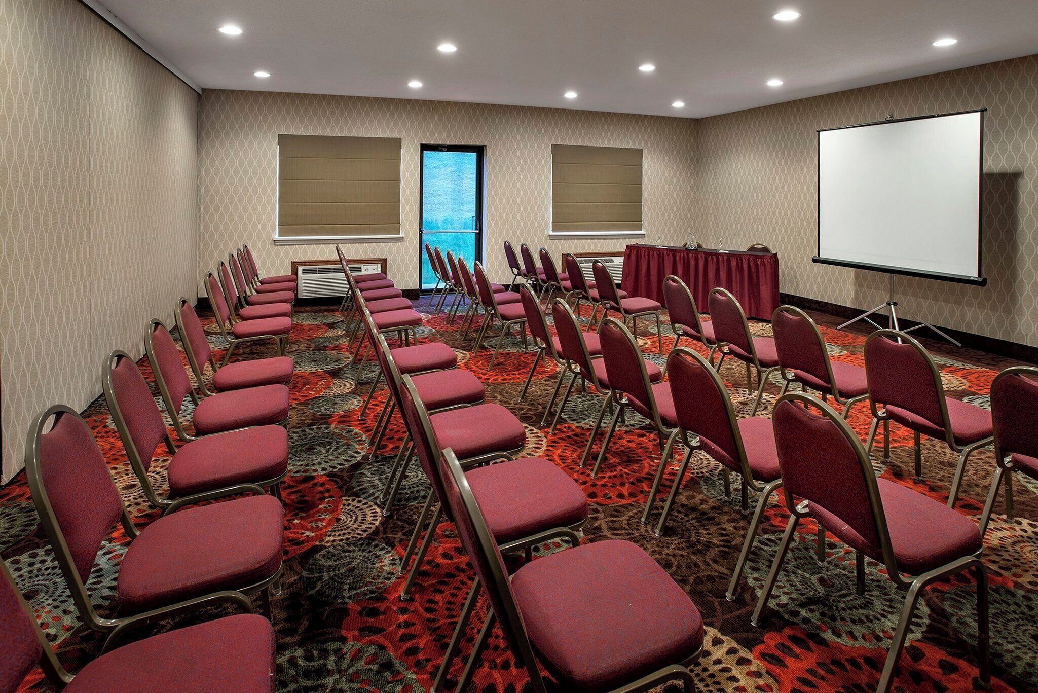 Uma sala de conferências com fileiras de cadeiras, uma tela de projetor e um tapete estampado.