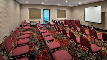 Uma sala de conferências com fileiras de cadeiras, uma tela de projetor e um tapete estampado.