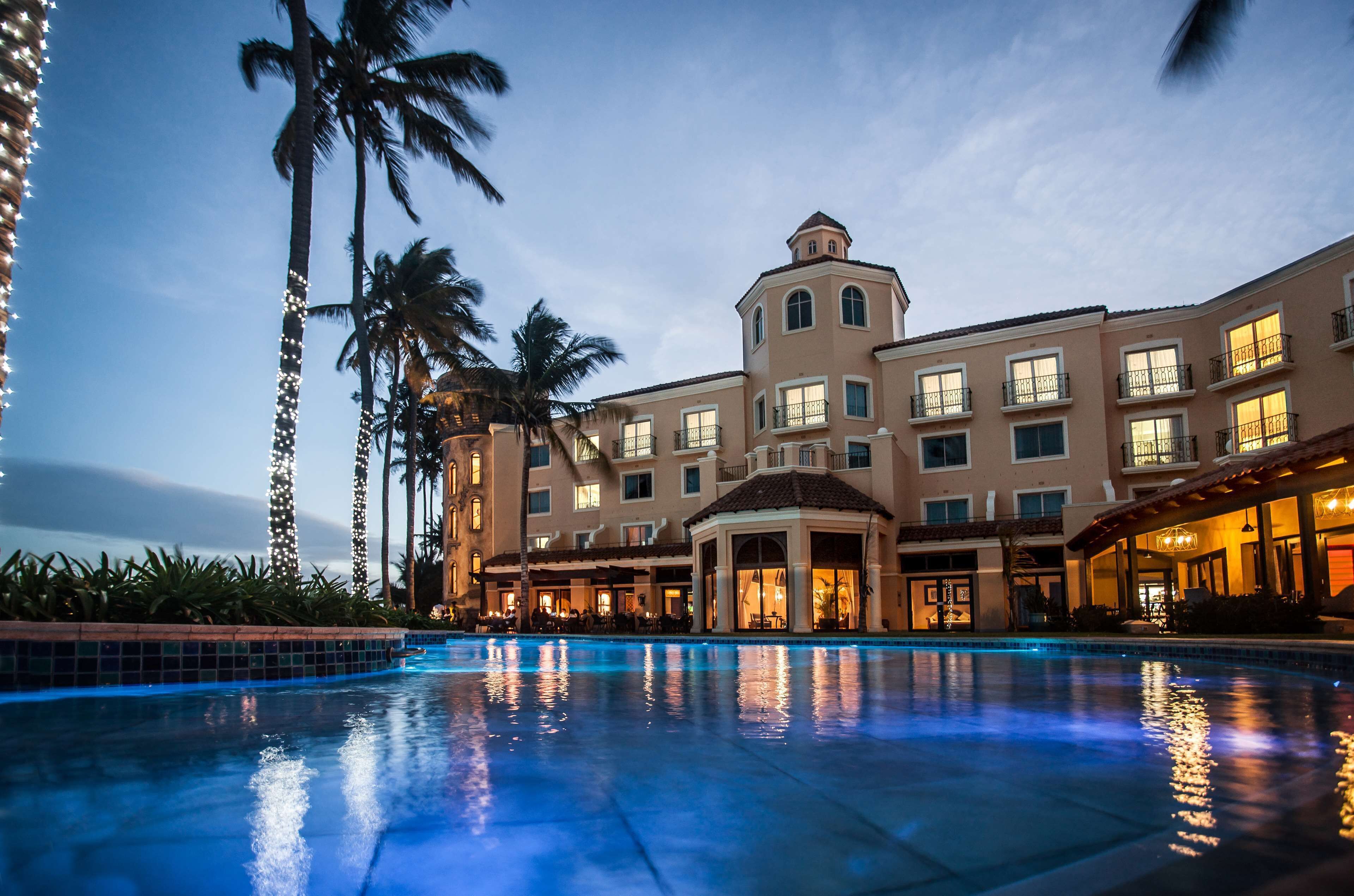 Um hotel com uma piscina grande, palmeiras e fachada bem iluminada.