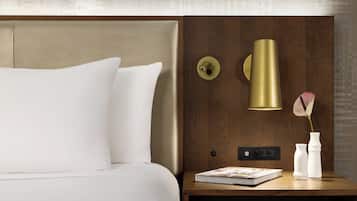 Uma cama bem arrumada com lençóis brancos, um criado-mudo de madeira com uma luminária e um vaso com uma única flor.