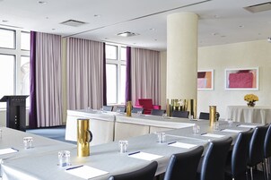 Uma sala de conferências com uma mesa comprida, cadeiras, um pódio e uma janela grande com cortinas.