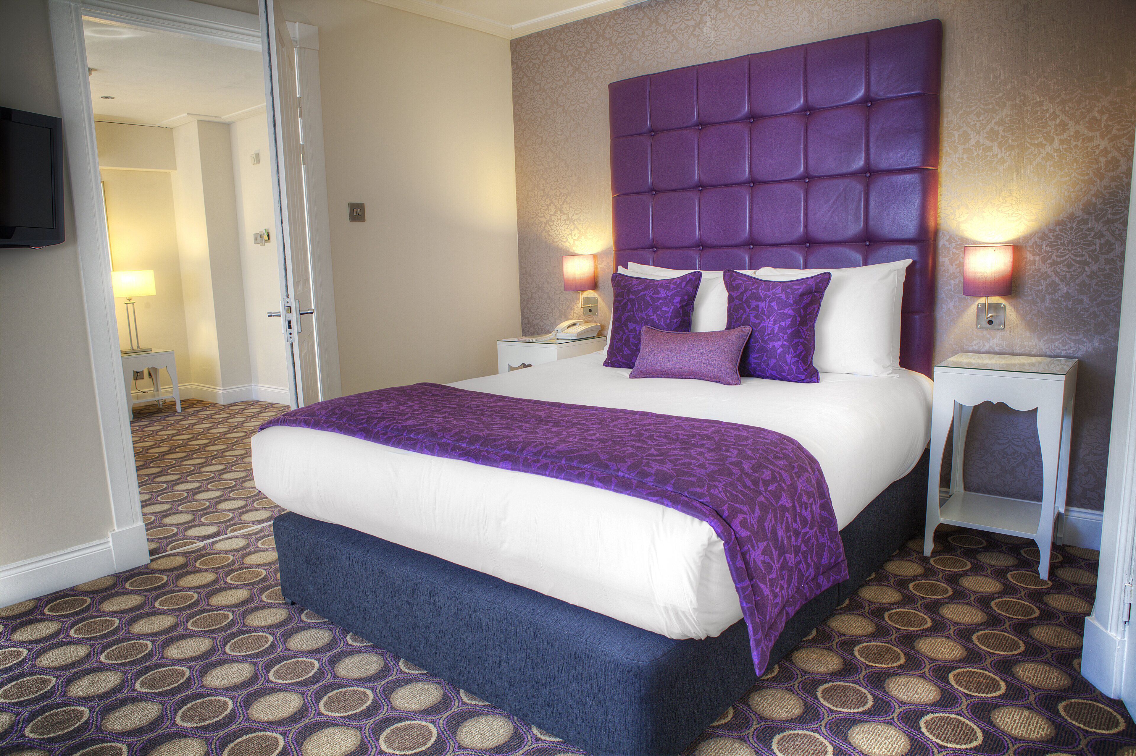 Quarto de hotel com uma cama grande, cabeceira roxa e carpete estampado.