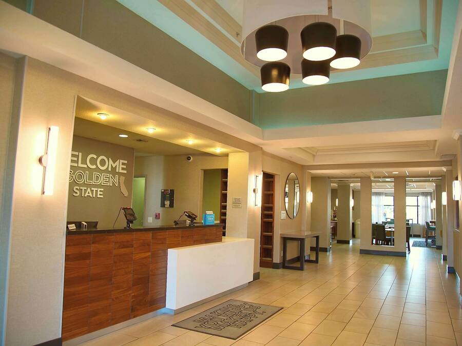 Uma área de recepção de hotel com balcão, um letreiro "Welcome Golden State", balcão de atendimento e um corredor que dá acesso a outros quartos.