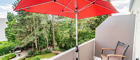 Uma varanda com um guarda-sol vermelho, cadeiras de vime e vista para um jardim com árvores e churrasqueira.