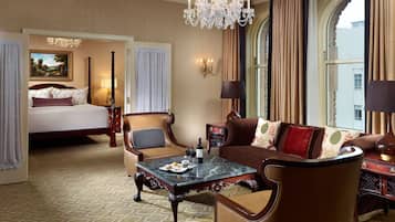 Quarto de hotel com cama, área de estar com sofá e poltronas, mesa de jantar e uma garrafa de vinho, além de um candelabro.