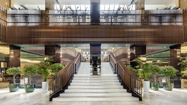 Um hall de hotel espaçoso e moderno, com uma escadaria central, grandes vasos de plantas e teto alto.