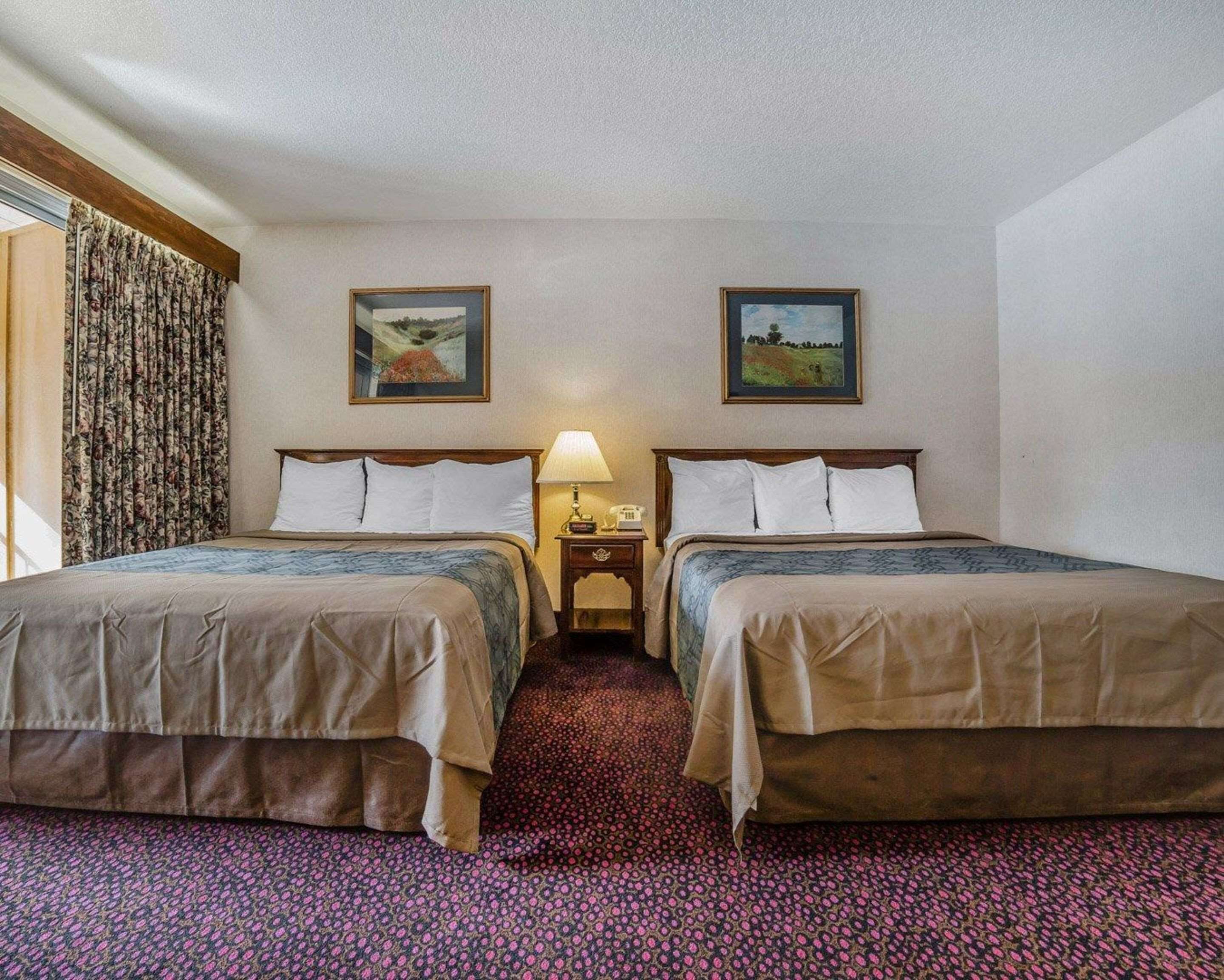 Quarto de hotel com duas camas, uma mesa de cabeceira com abajur e duas imagens emolduradas na parede.