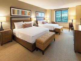 Quarto de hotel com duas camas, uma escrivaninha, uma cadeira e uma janela com vista para o verde.