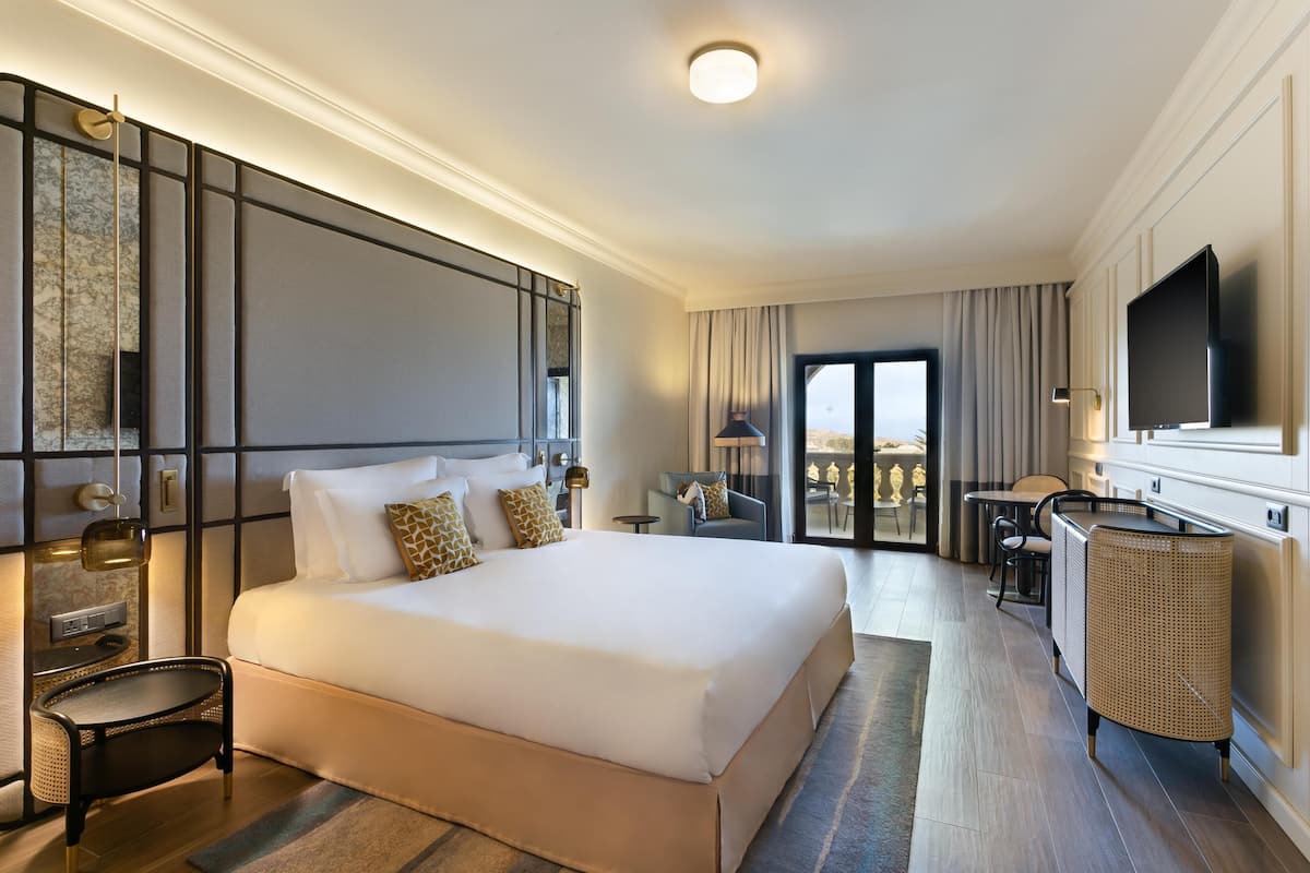 Quarto de hotel com uma cama grande, uma escrivaninha, uma cadeira, televisão e varanda com vista.