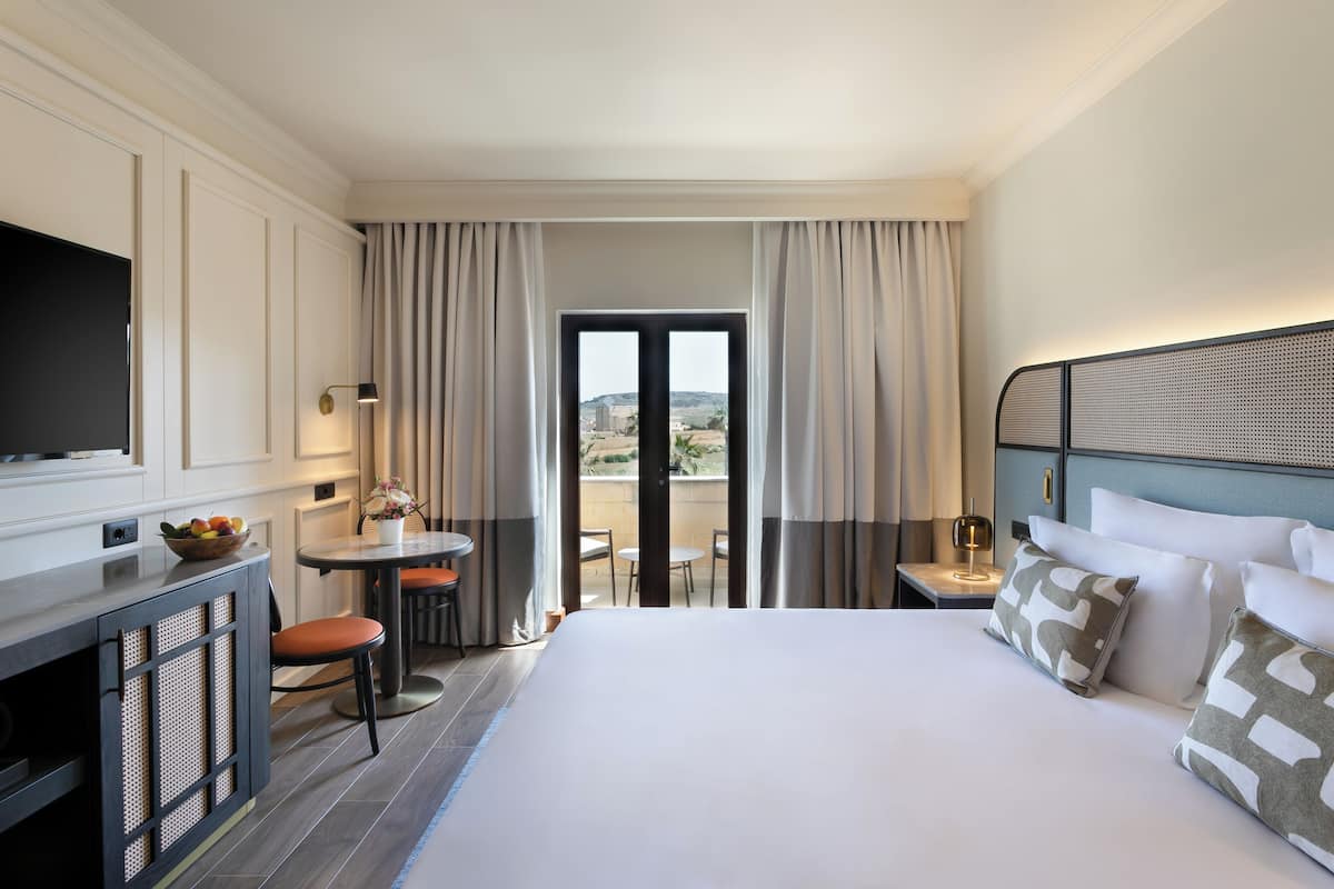 Quarto de hotel com cama, televisão, escrivaninha com cadeira, mesa pequena com cadeira e varanda com cortinas.