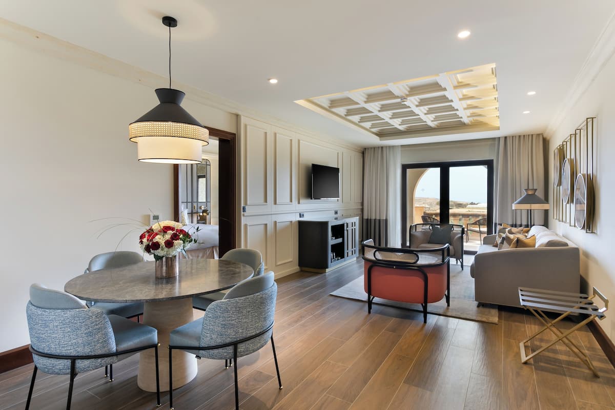 Quarto de hotel moderno com área de refeições, sofá, televisão e uma grande janela.