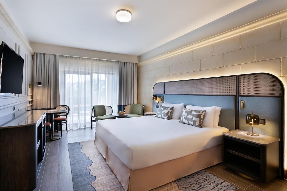 Quarto de hotel com uma cama grande, uma escrivaninha, uma cadeira, uma televisão e uma janela com cortinas.