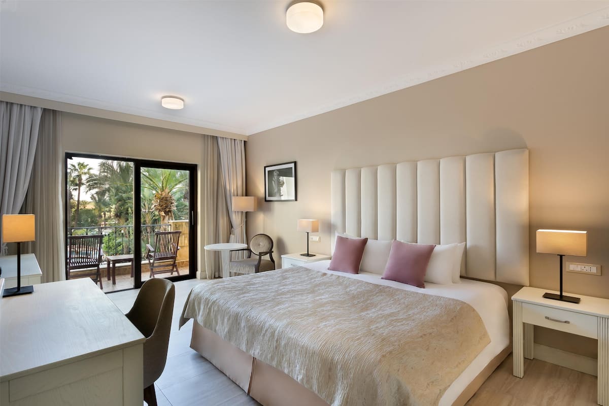 Quarto de hotel com uma cama grande, uma escrivaninha, uma cadeira, uma mesinha e uma varanda com vista para palmeiras.