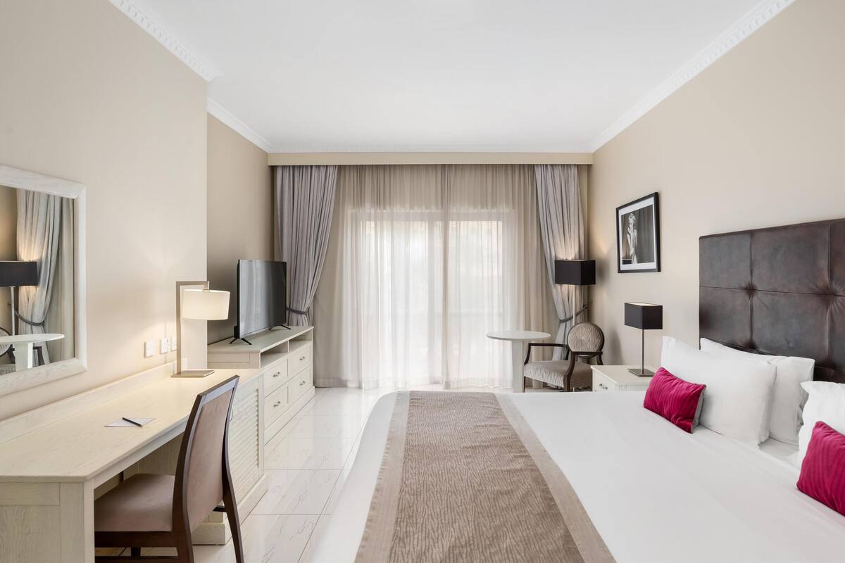 Quarto de hotel com cama, escrivaninha, cadeira, televisão e cortinas.