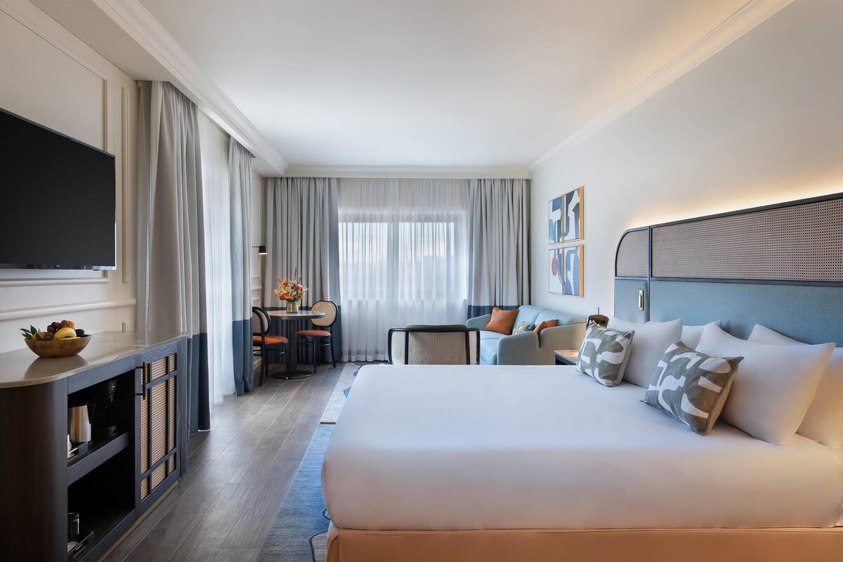 Quarto de hotel com uma cama grande, uma escrivaninha com cadeira, uma televisão e um espaço de refeições com mesa e cadeiras.