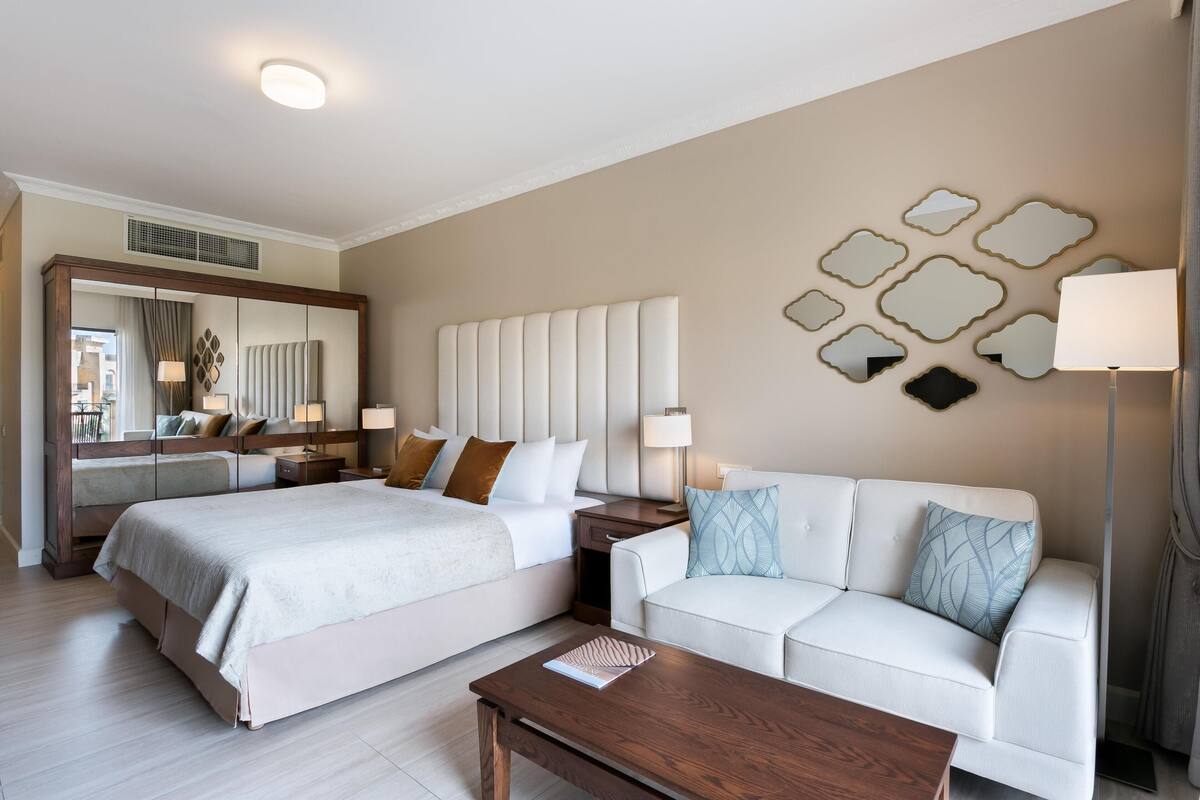 Quarto de hotel com uma cama grande, um sofá, uma mesa de centro e uma parede com espelhos decorativos e luminárias.