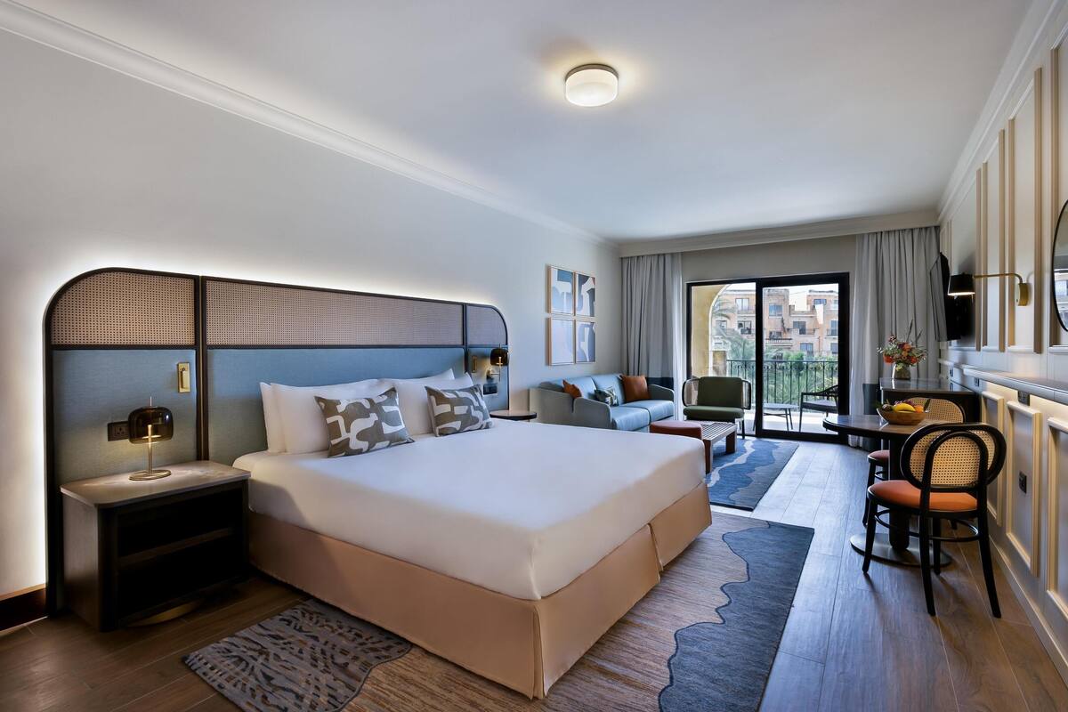 Quarto de hotel moderno com uma cama grande, área de estar com sofá e poltronas, mesa de jantar e vista da cidade através de amplas janelas.