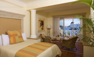Quarto de hotel com cama, uma cadeira, uma mesinha com frutas, uma luminária e vista para o mar e edifícios.