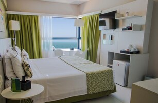Quarto de hotel com cama, escrivaninha, cadeira, televisão e vista para o mar.