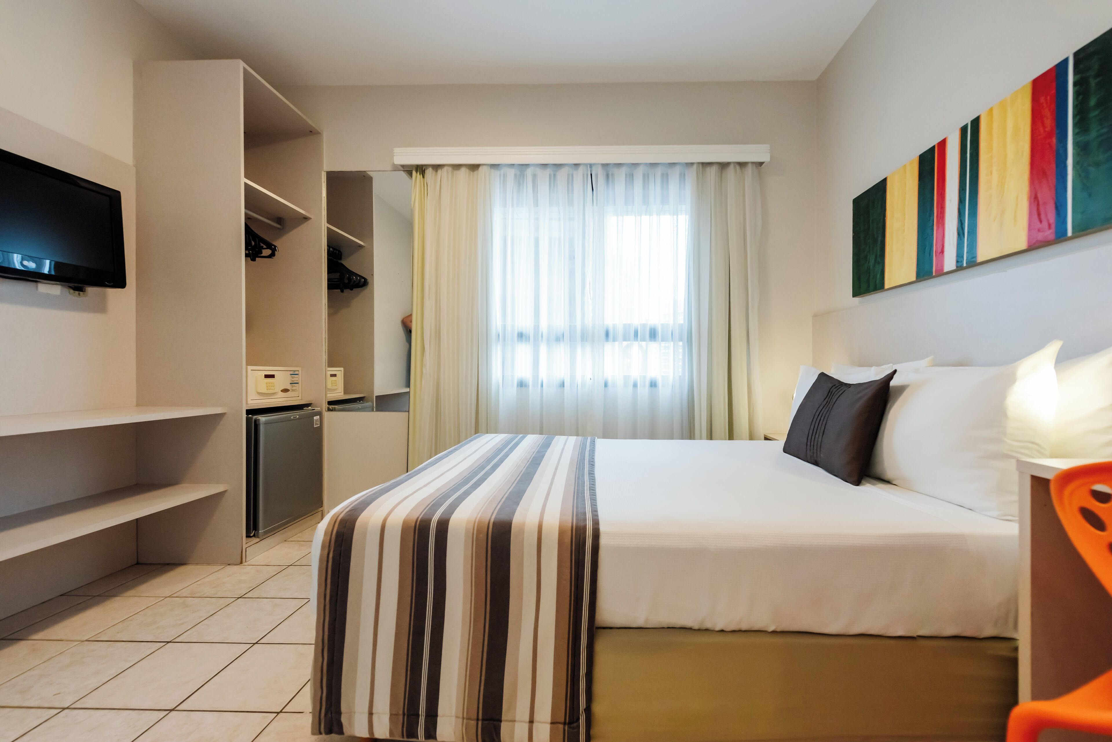 Quarto de hotel com uma cama, televisão na parede, secretária com computador e janela com cortinas.